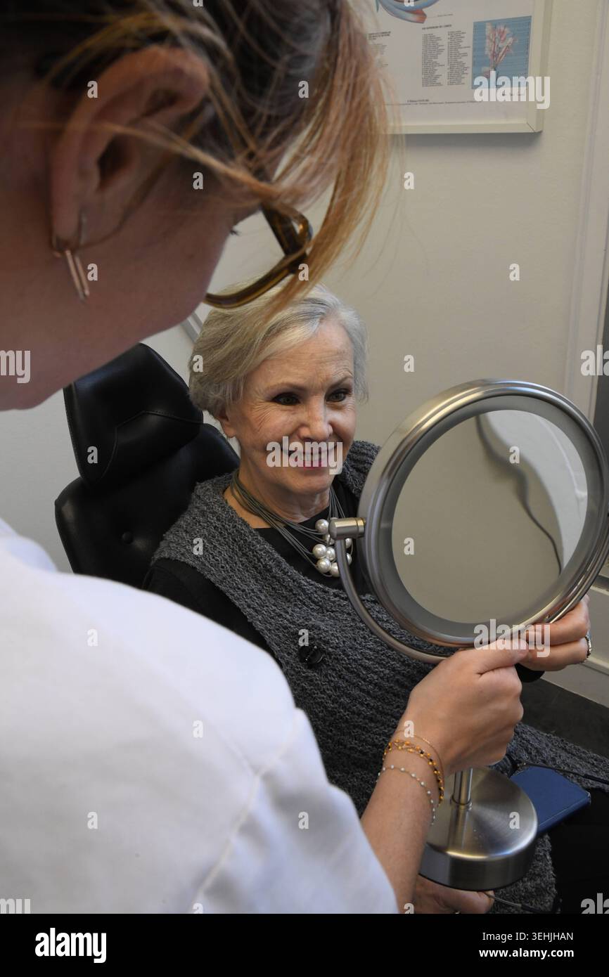 A person in a white coat holds a mirror for a smiling woman sitting on ...