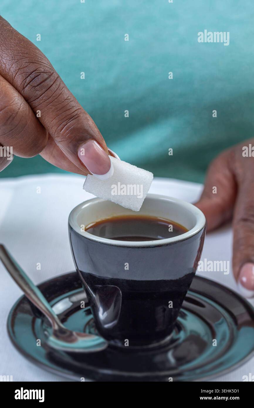 A hand holds a white sugar cube over a small black cup of coffee. The ...