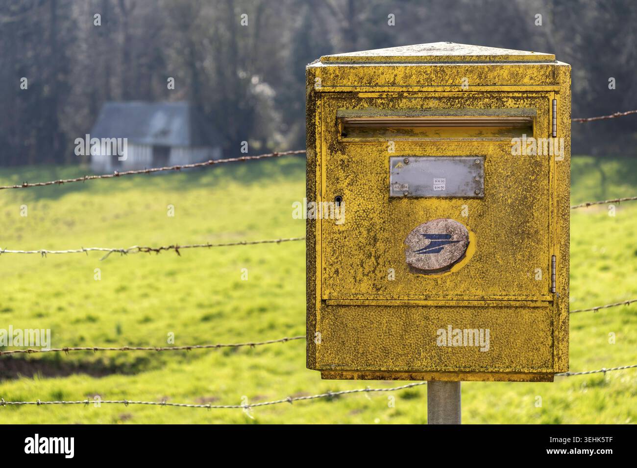 A weathered yellow mailbox stands on a metal post behind a barbed wire ...