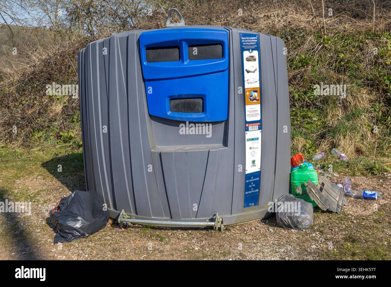 A grey and blue recycling bin stands outdoors, surrounded by black ...