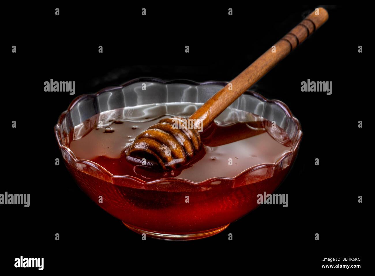 A glass bowl contains a viscous amber liquid and a wooden honey dipper ...