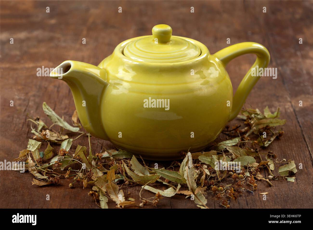 A yellow teapot with a lid sits on a wooden surface. Dried leaves and ...