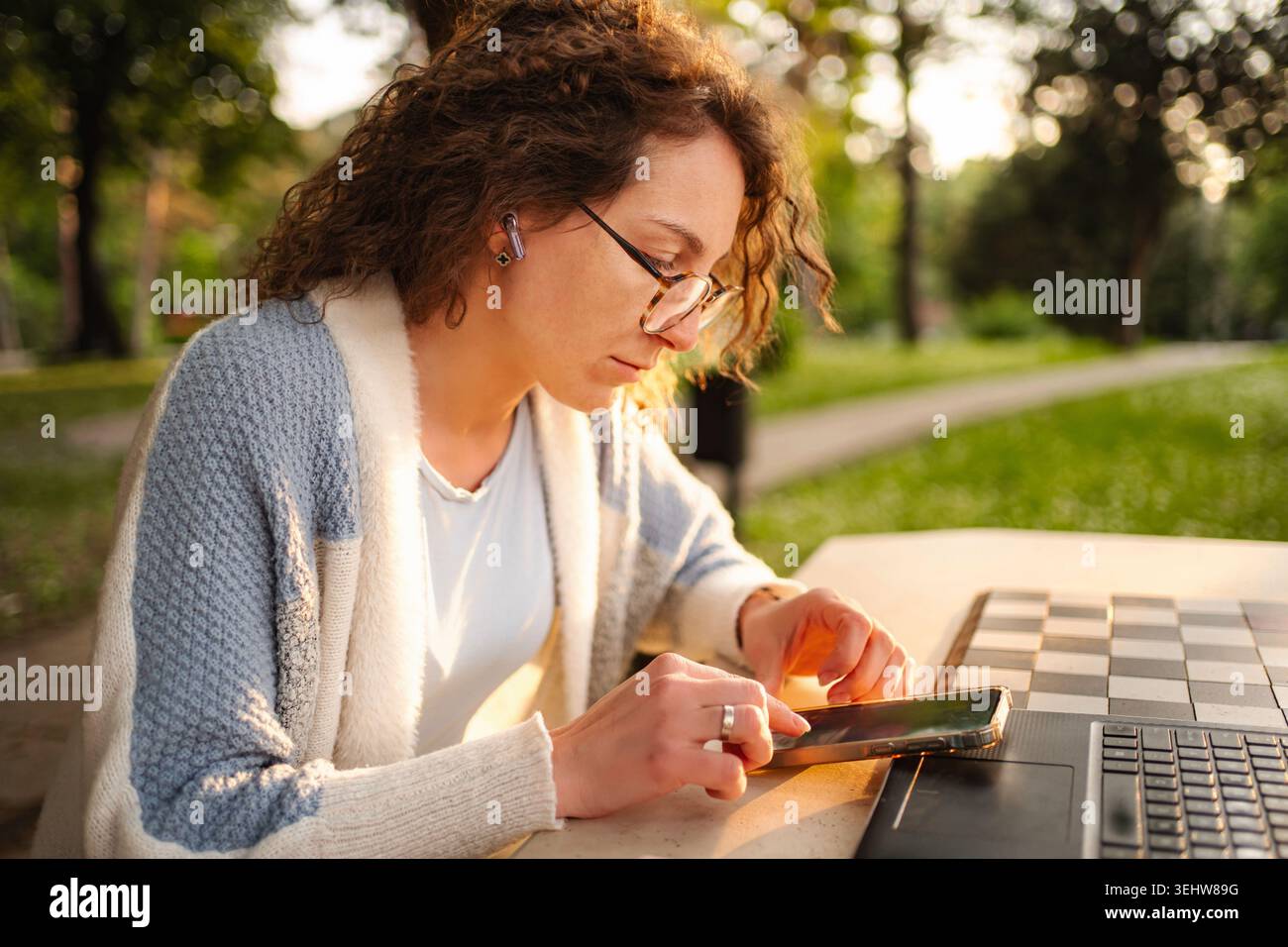 Young adult woman using smartphone and laptop for remote work outdoors ...
