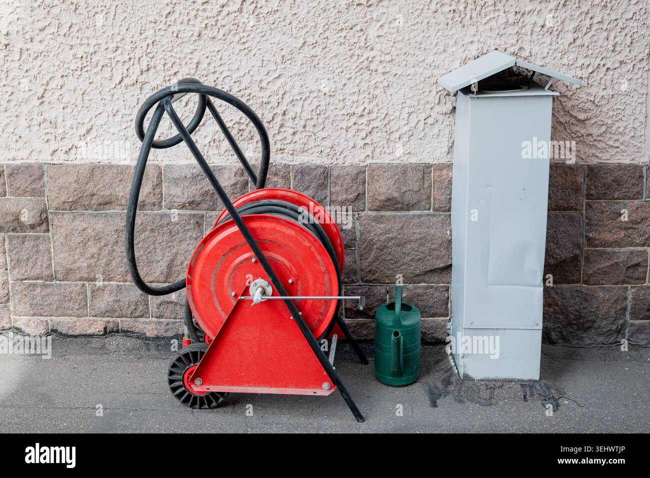 Red fire hose reel with watering can near textured residential wall in ...