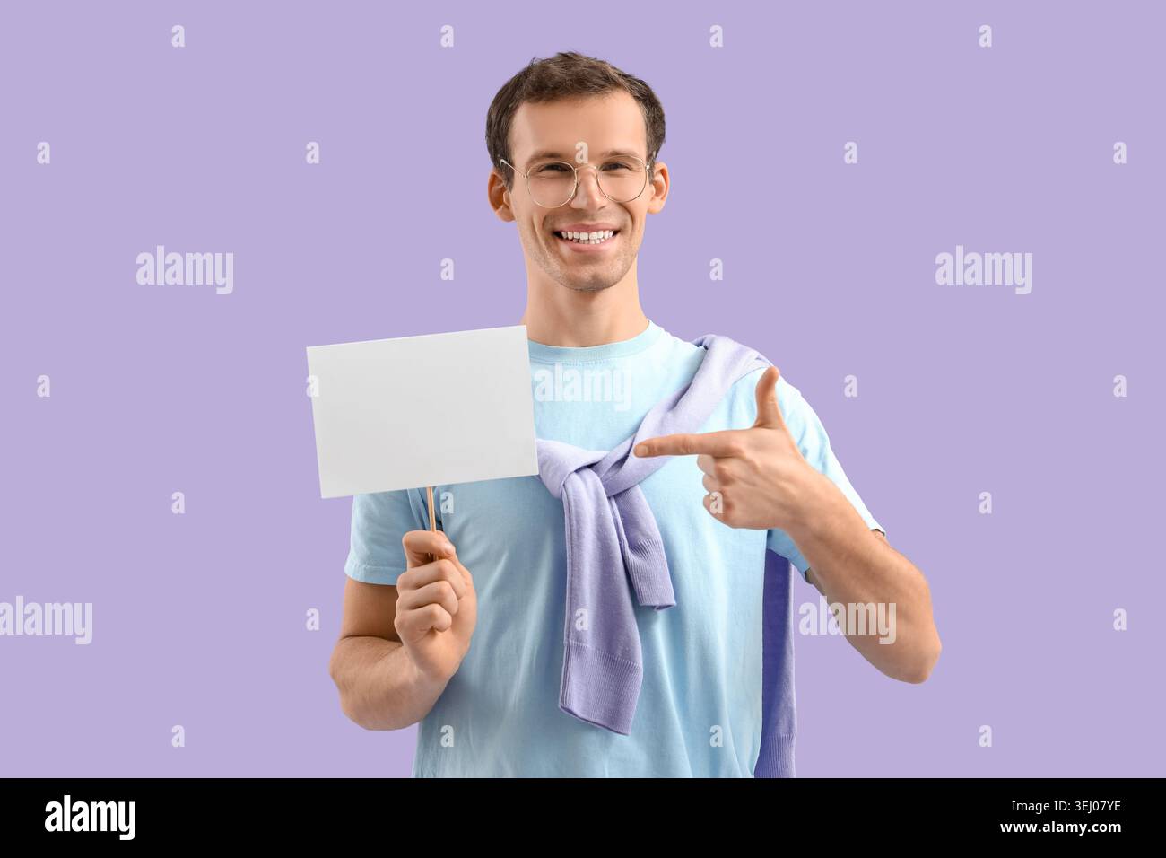 Young man pointing at blank paper sign on lilac background Stock Photo ...