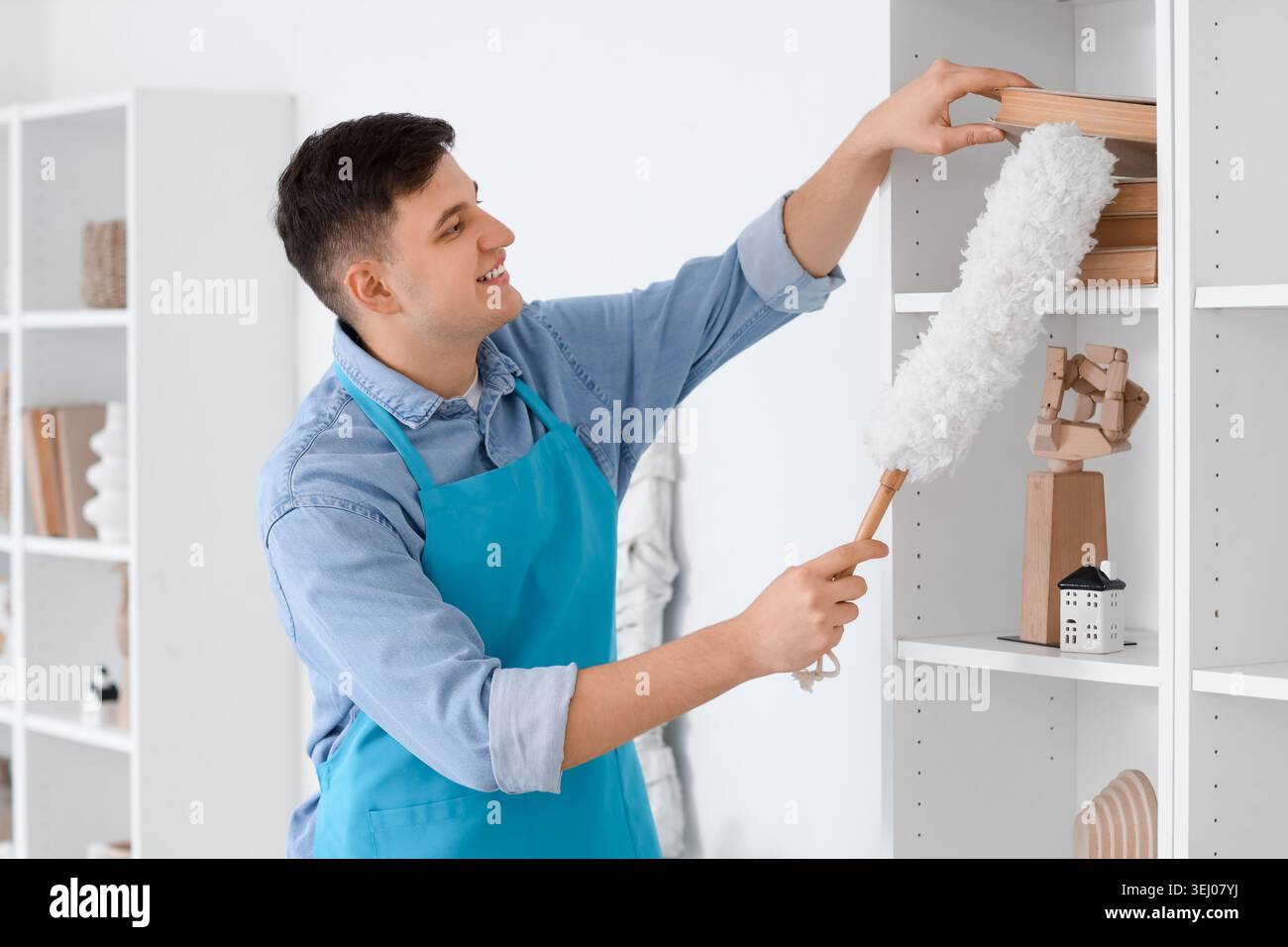 Young man in apron cleaning shelf with pp-duster at home Stock Photo ...