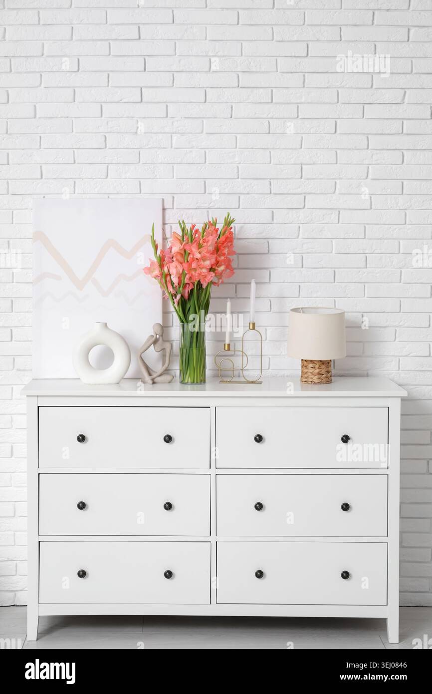 Vase with gladiolus flowers, lamp and decor on commode near white brick ...