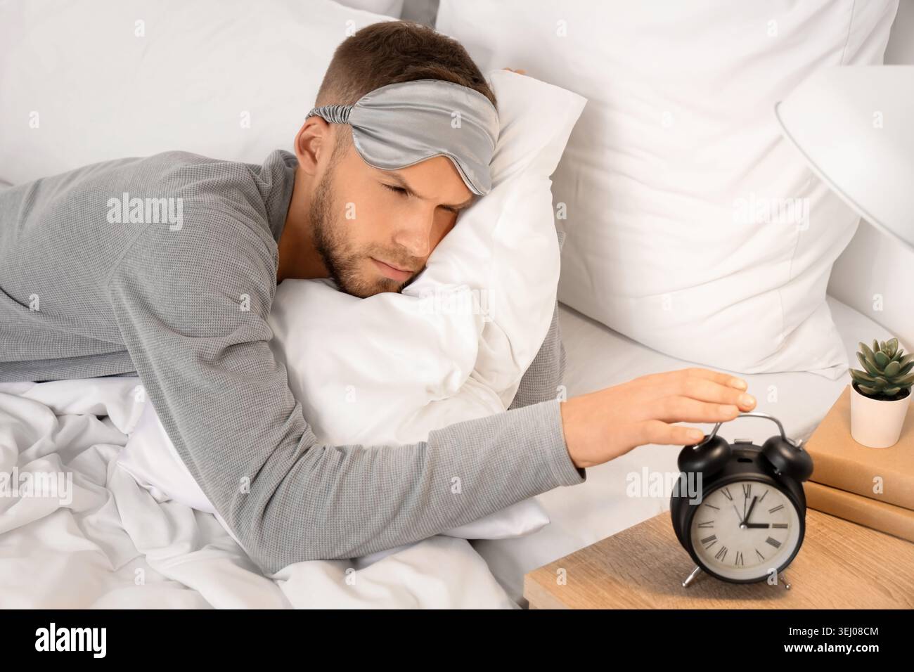 Young man in sleeping mask turning off alarm clock while lying on bed ...