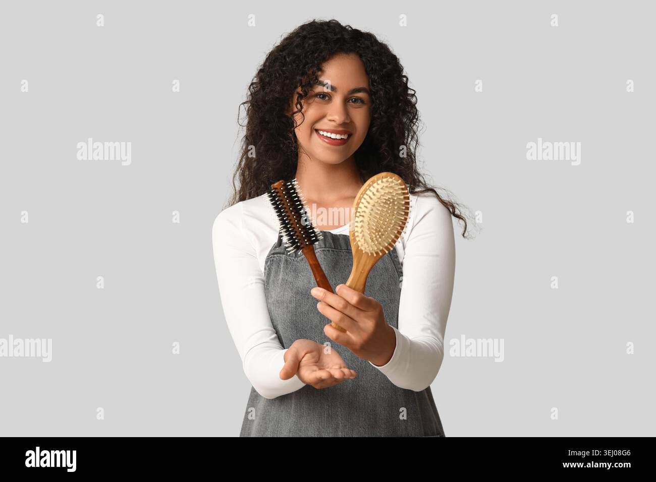 Beautiful young happy African-American hairdresser with brushes on grey ...