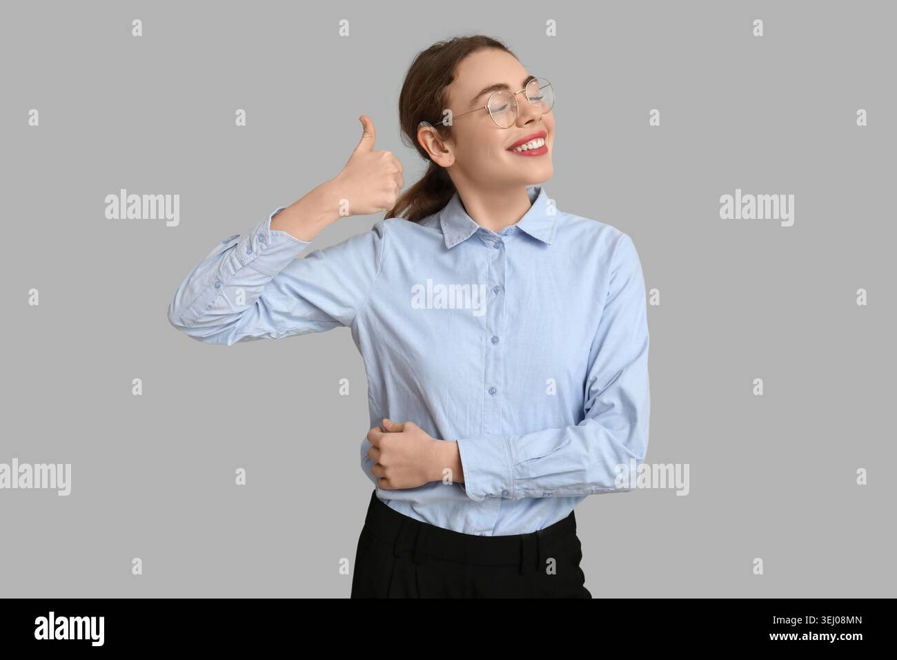 Beautiful young woman with hearing aid showing thumb-up gesture on grey ...
