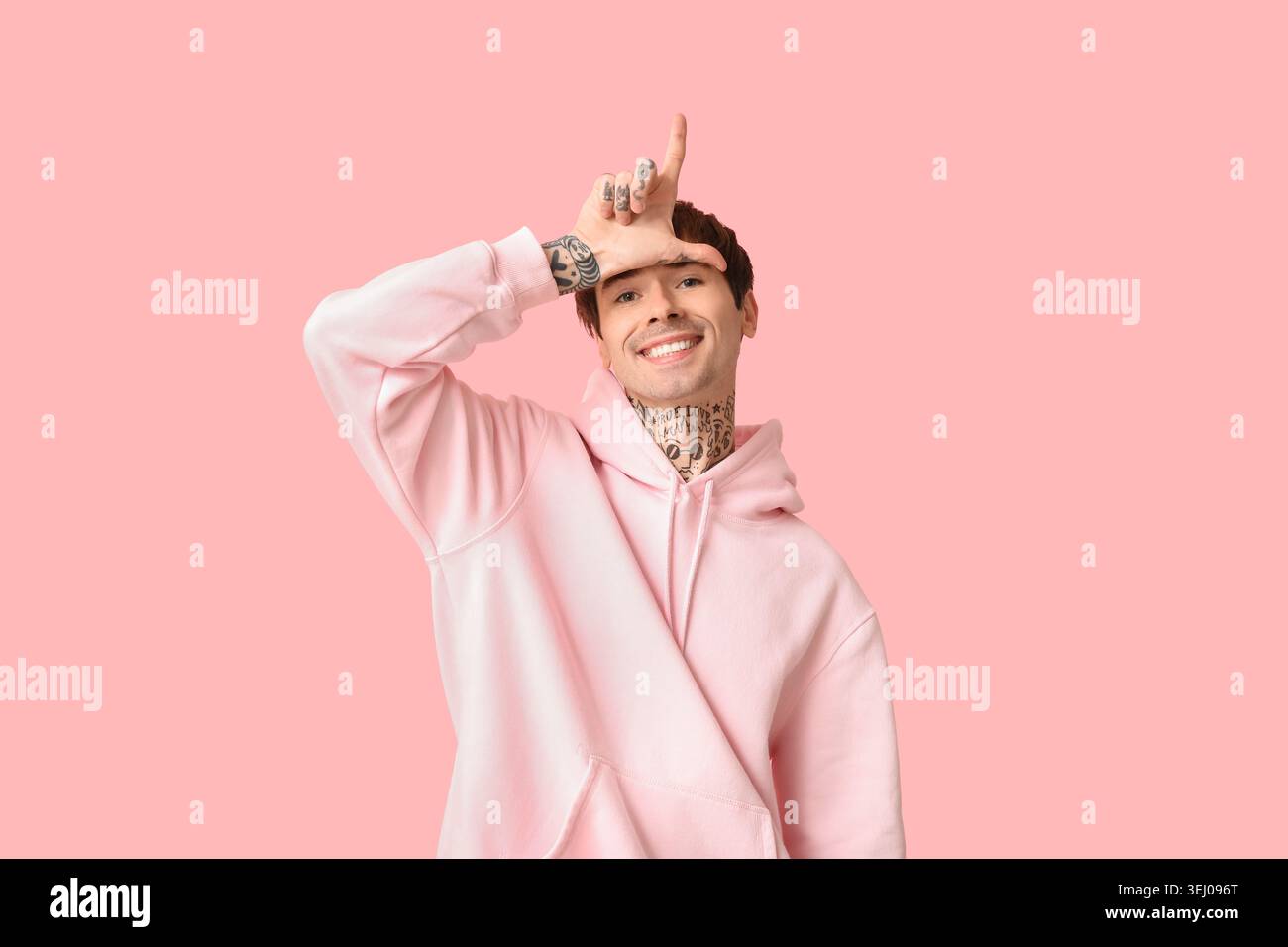 Happy handsome young tattooed man showing loser gesture on pink ...