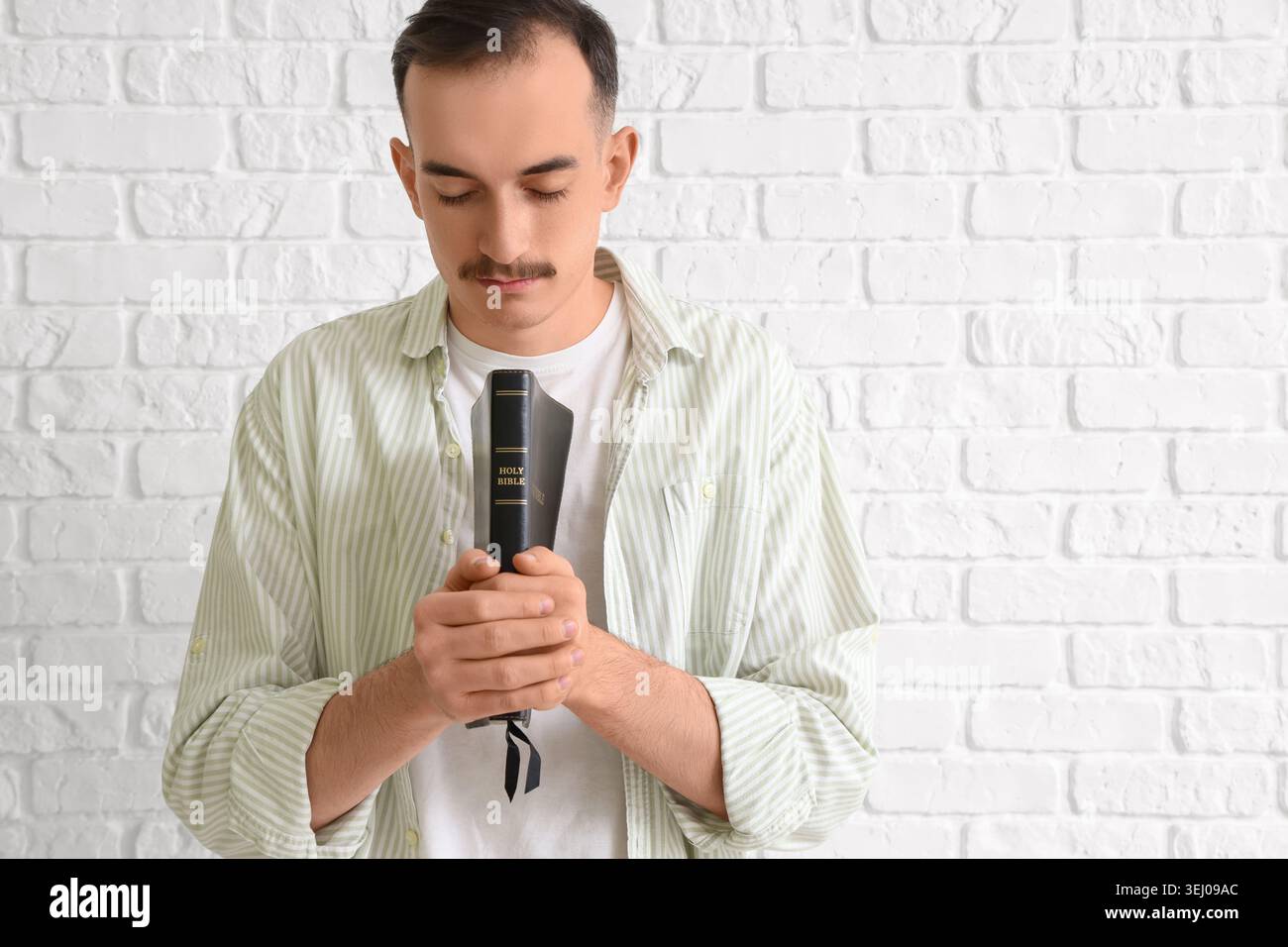 Young man with Holy Bible praying on white brick background Stock Photo ...