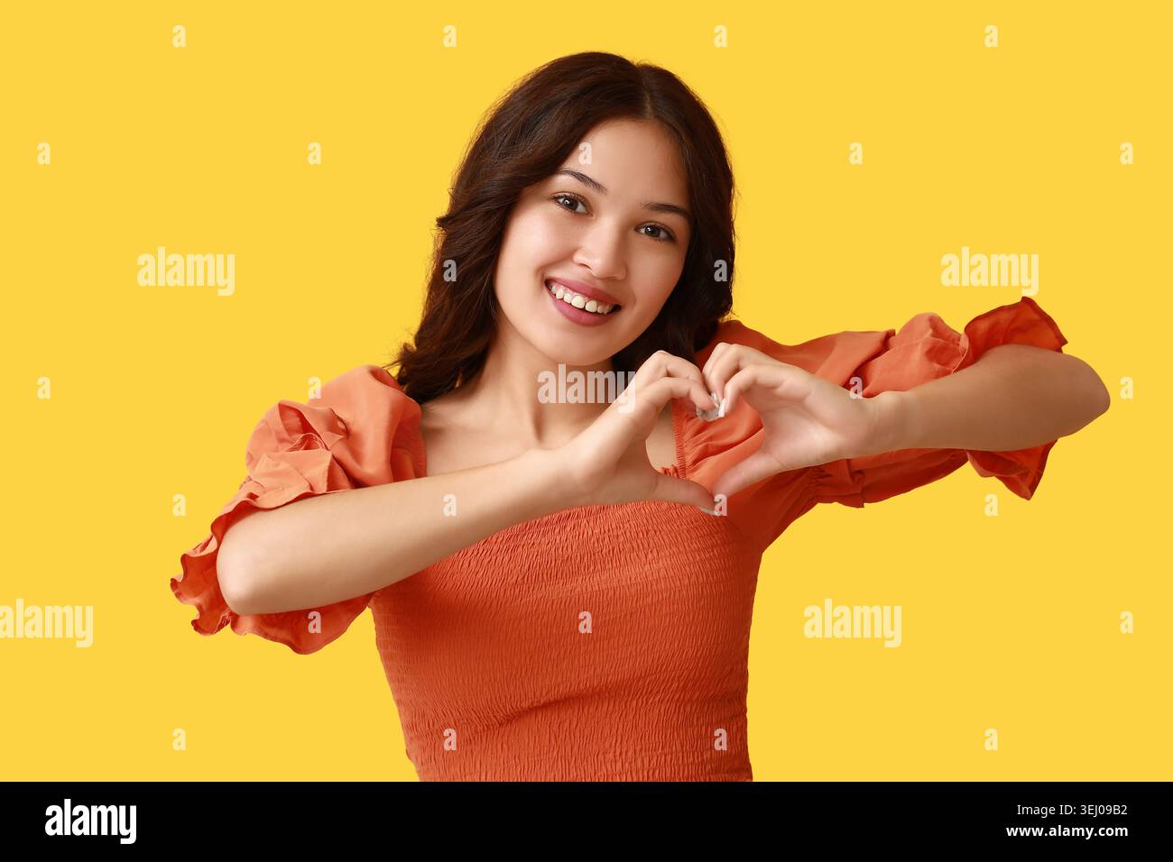 Happy young Asian woman showing heart gesture on yellow background ...