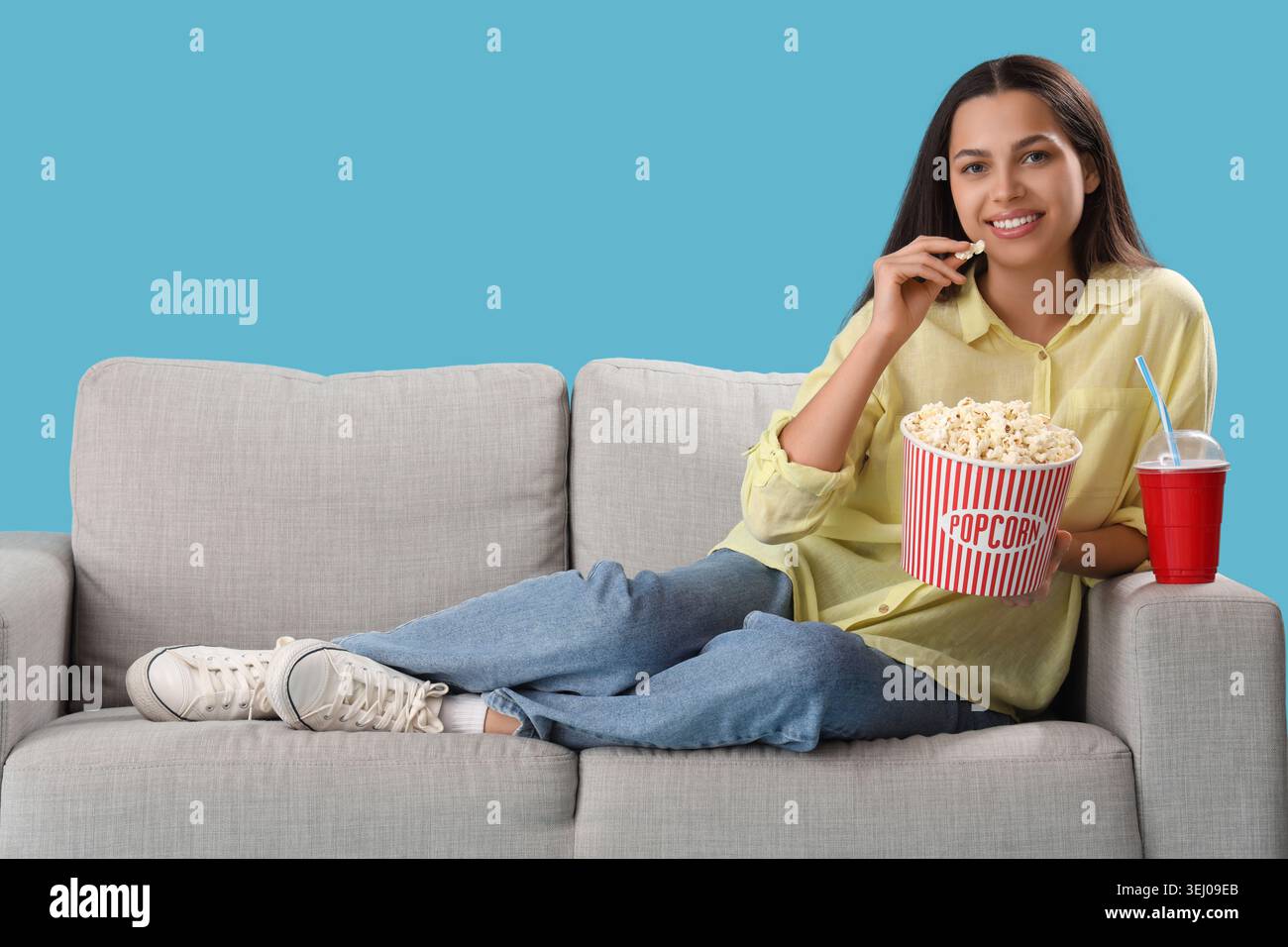 Young woman with popcorn watching movie on sofa against blue background ...