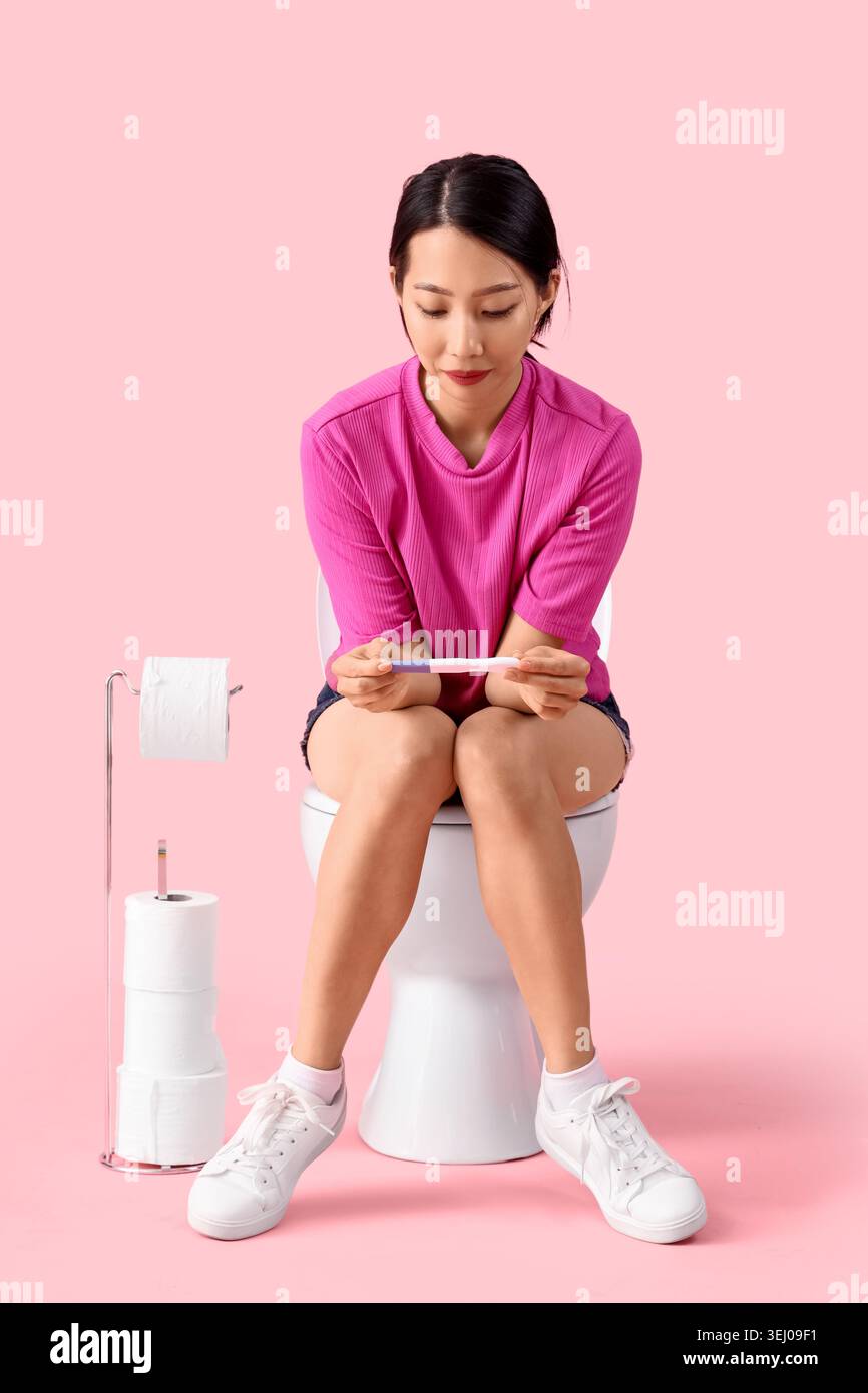 Young Asian woman with pregnancy test sitting on toilet bowl against ...