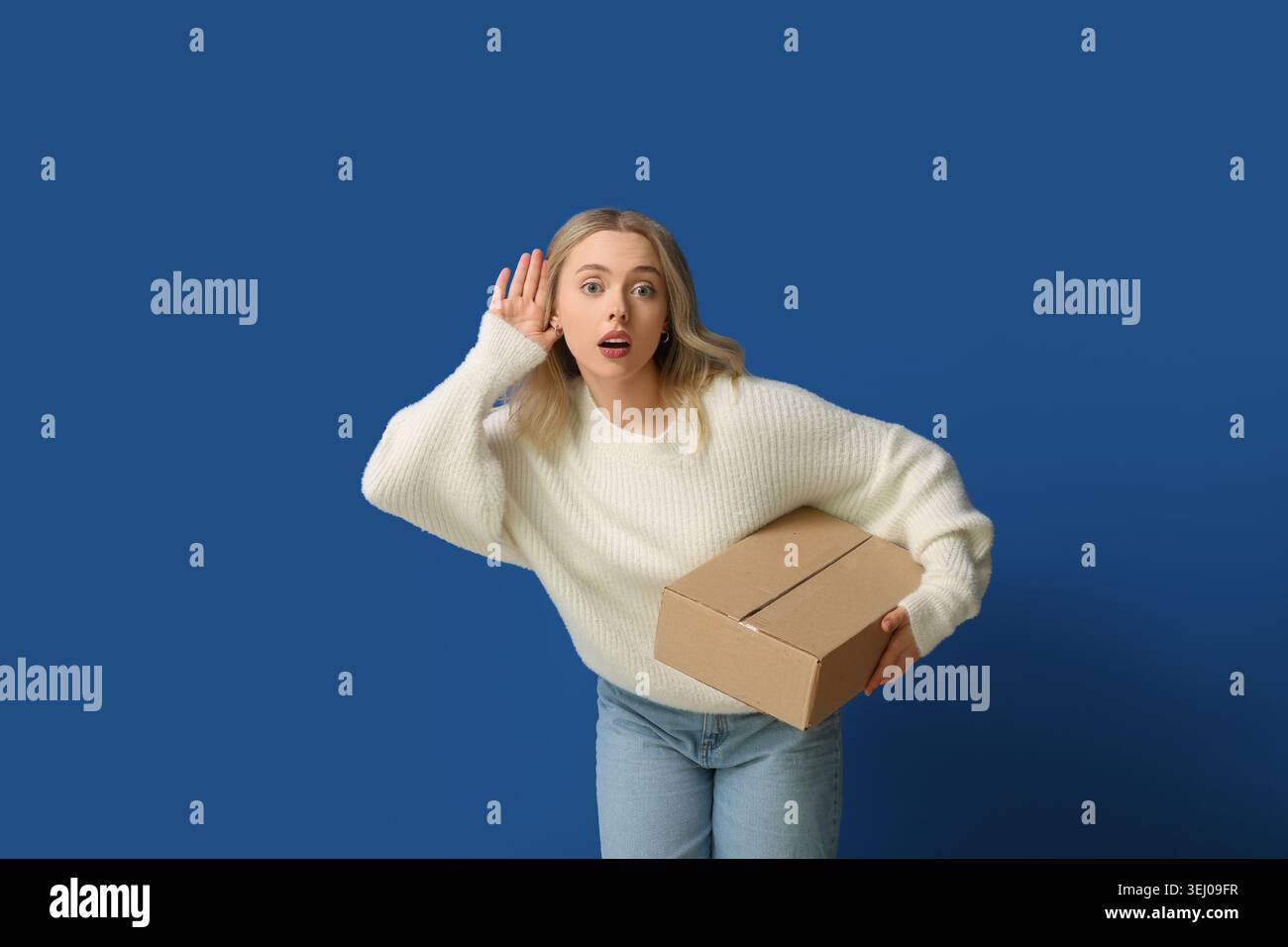 Young woman with cardboard box trying to hear something on blue ...