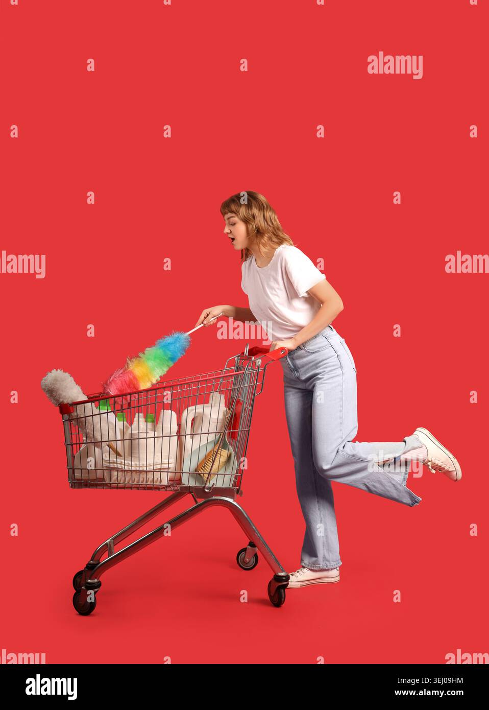 Young woman with cleaning supplies in shopping cart on red background ...