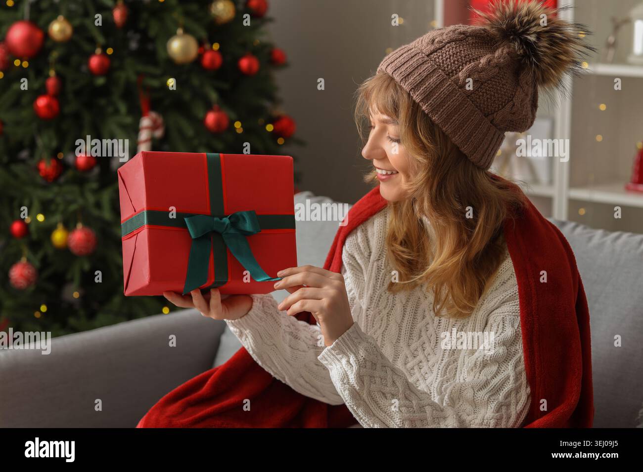 Pretty young woman with Christmas gift box sitting on sofa at home ...