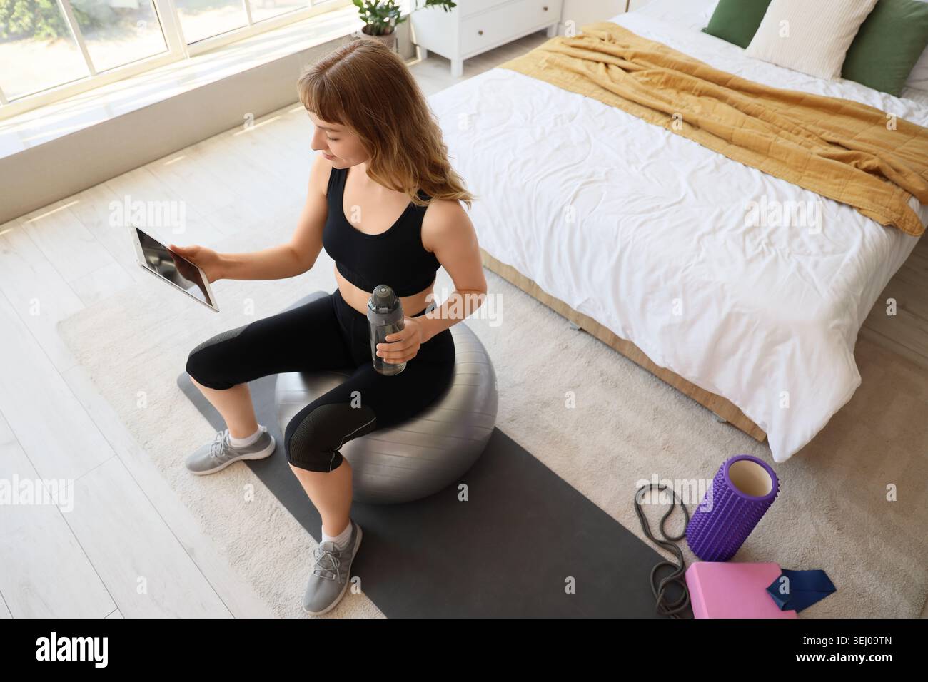 Young sporty woman with bottle of water and tablet sitting on fitness ...
