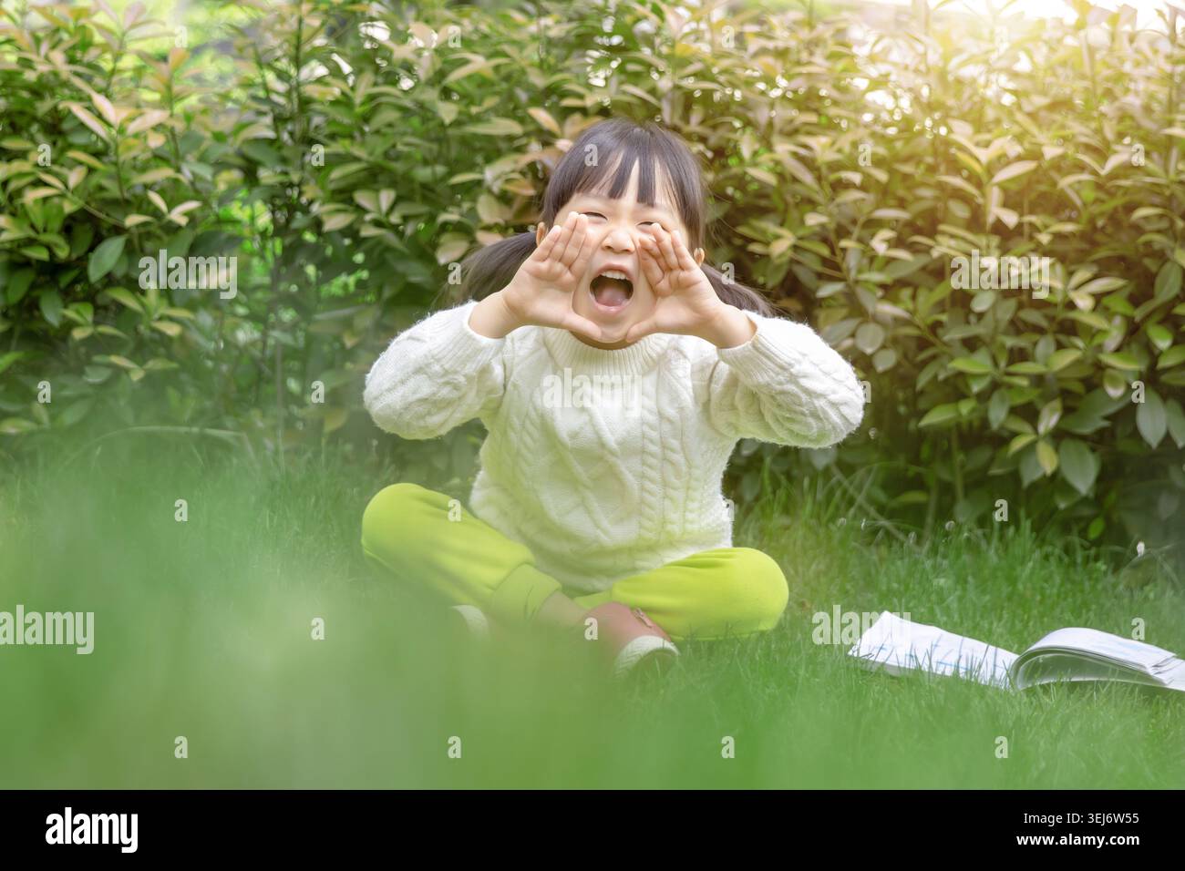 A cute Asian girl sits on the grass outdoors, making a shouting gesture ...