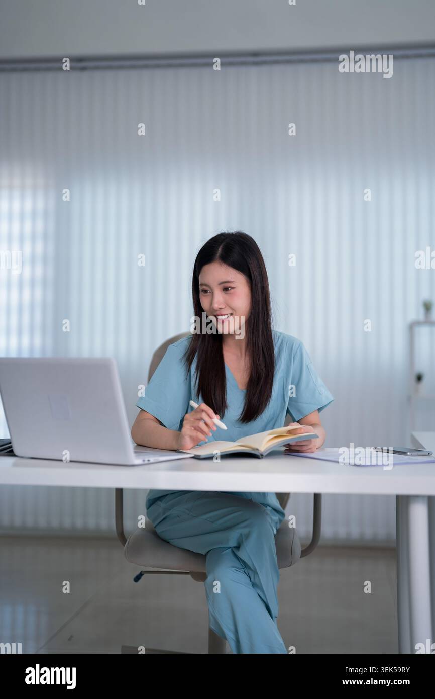 Young Asian female doctor in scrubs writing notes while using laptop ...