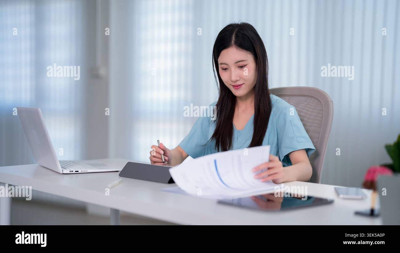 Asian woman in blue scrubs reviewing documents at desk with laptop ...