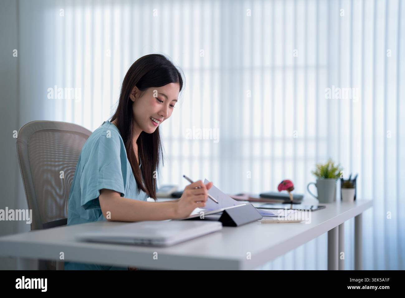 Young Asian woman sitting at desk writing notes on tablet smiling ...