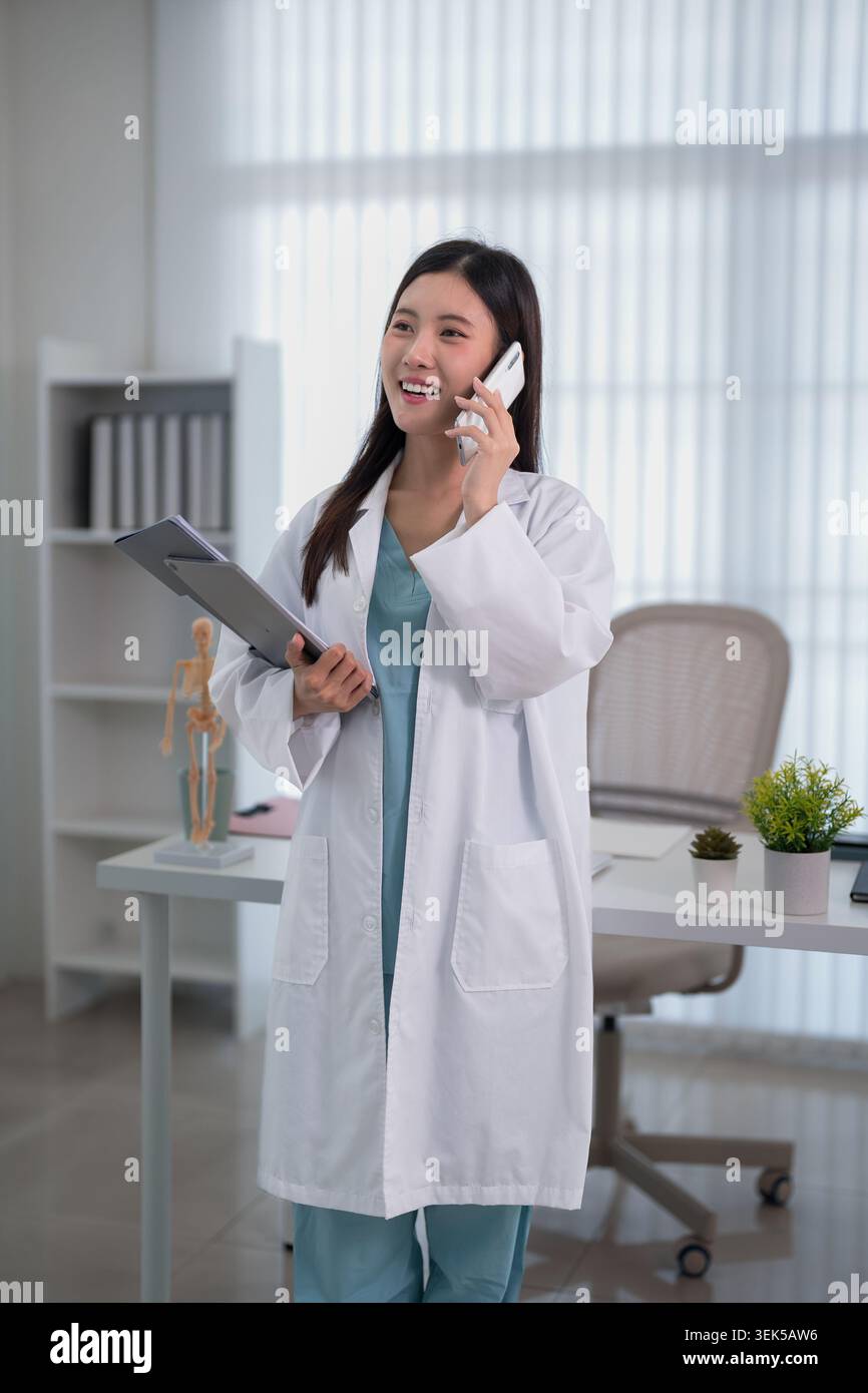 Asian female doctor in white coat talking on phone while holding tablet ...