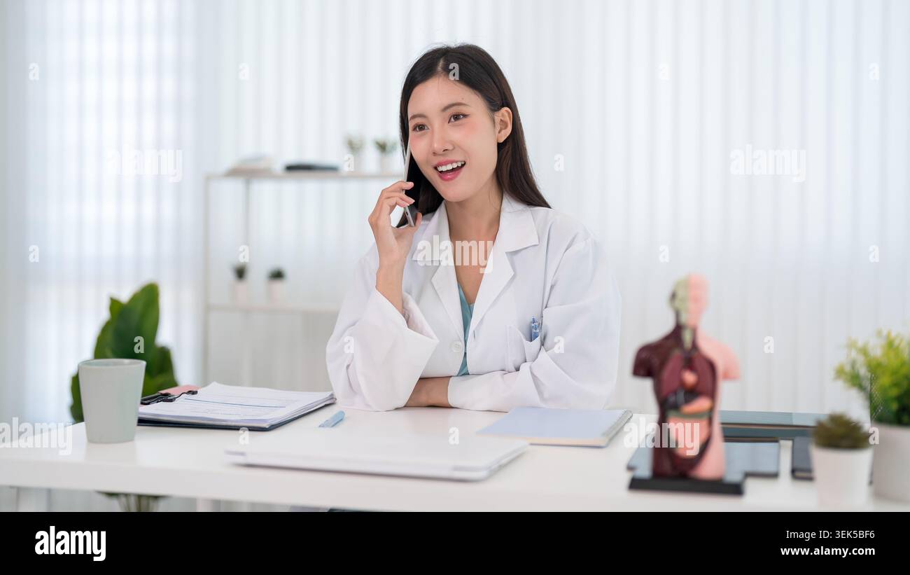 young Asian doctor speaking on phone at clinic desk providing medical ...