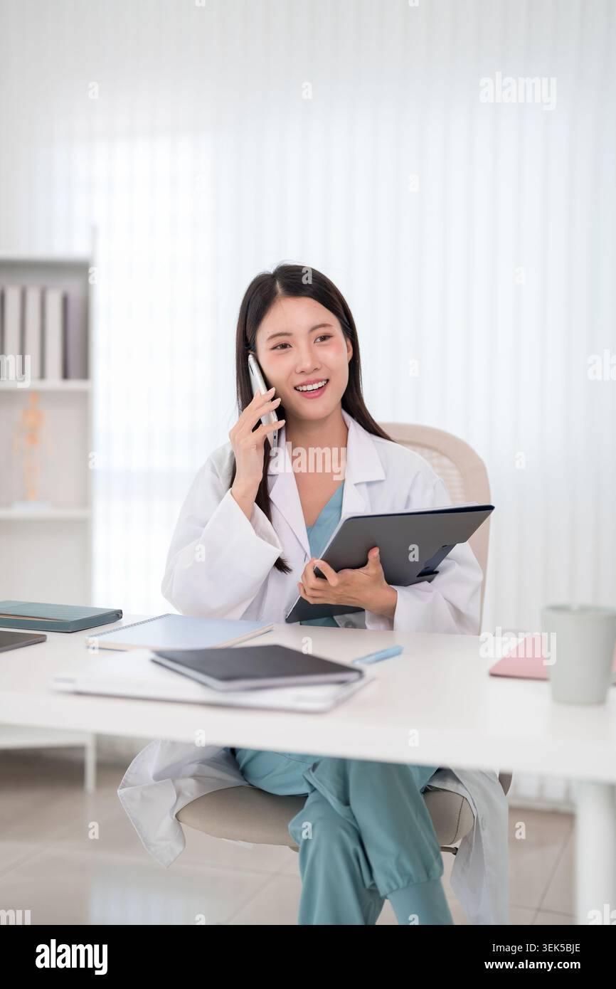 Asian female doctor in white coat talking phone holding tablet modern ...