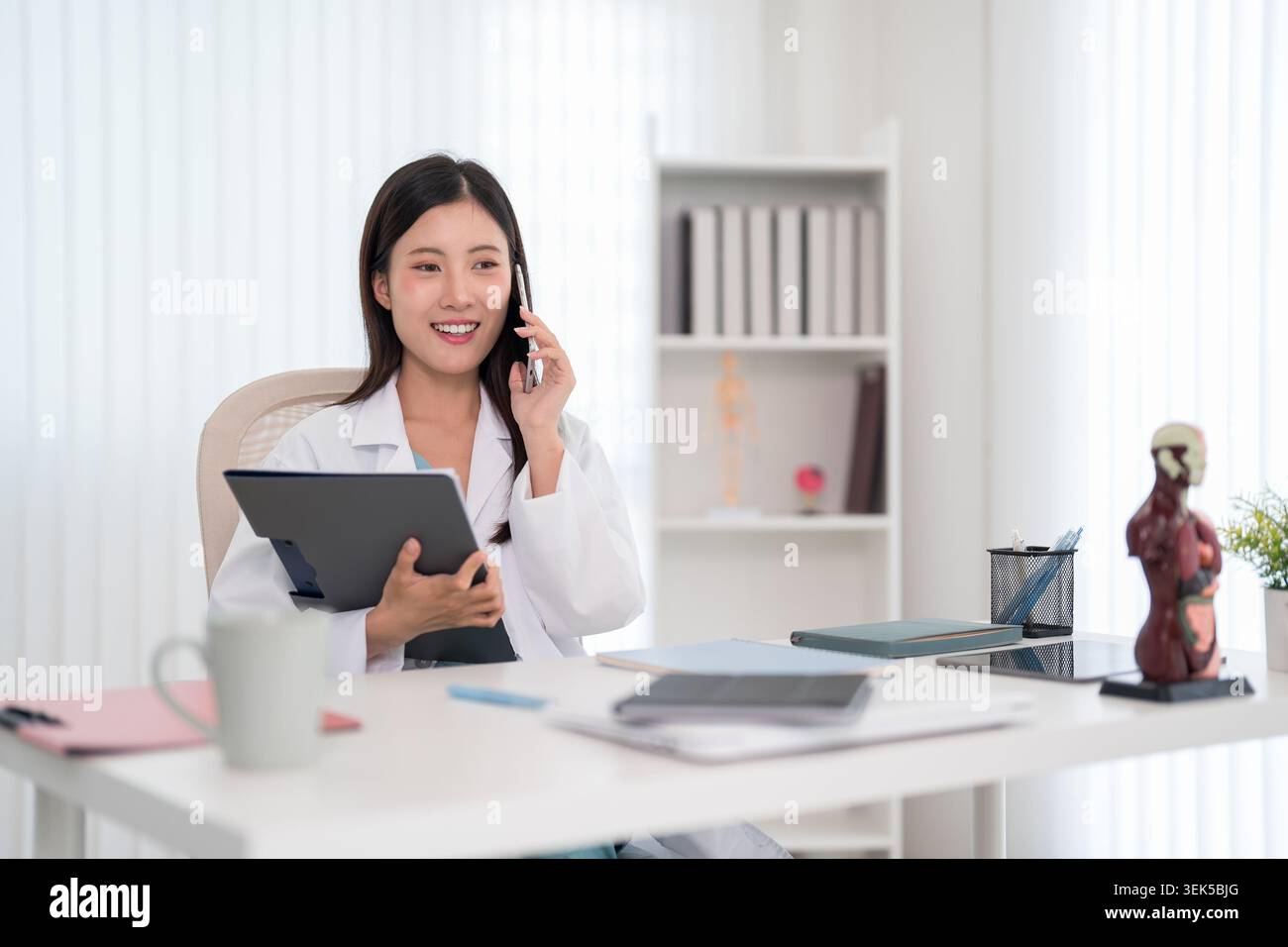 Young Asian woman doctor in white coat talking on mobile phone holding ...