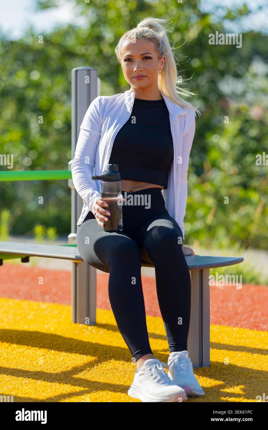 A young woman in fitness attire relaxes with a water bottle on a sunny ...