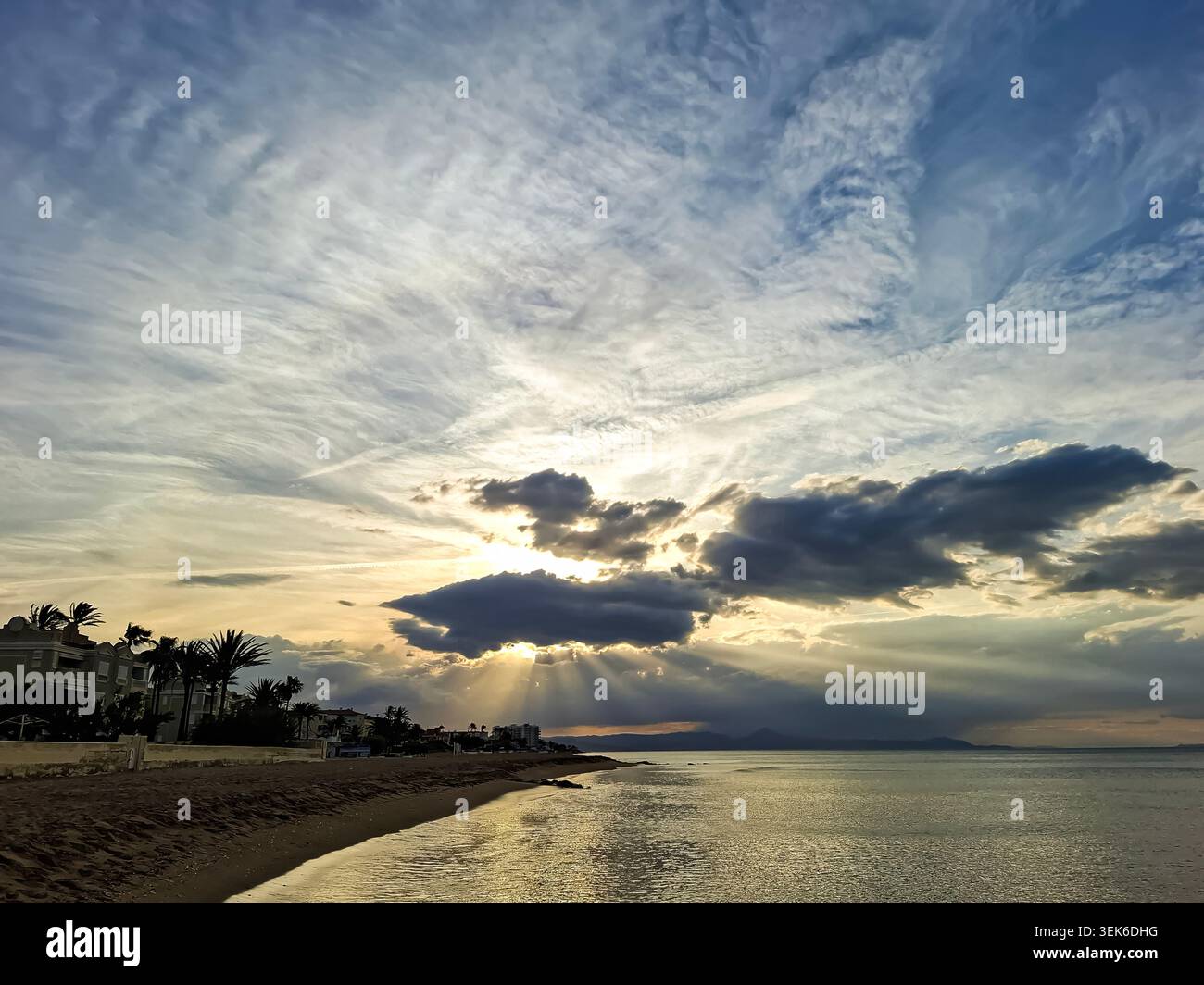 The sun sets over the beach, casting light through dark clouds ...