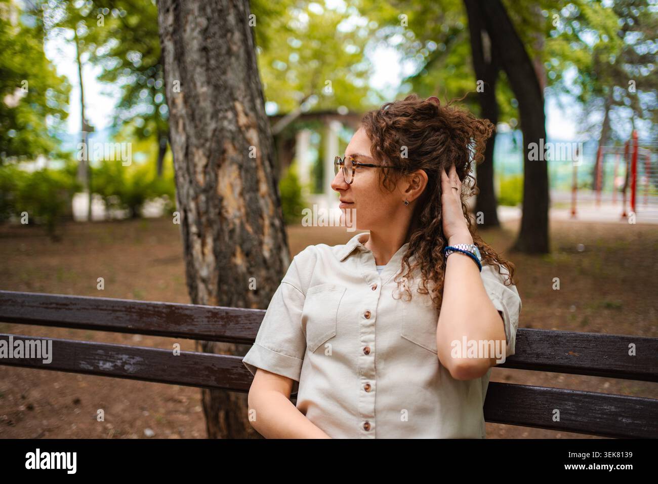 Woman with curly hair and glasses sitting on a park bench, reflecting ...