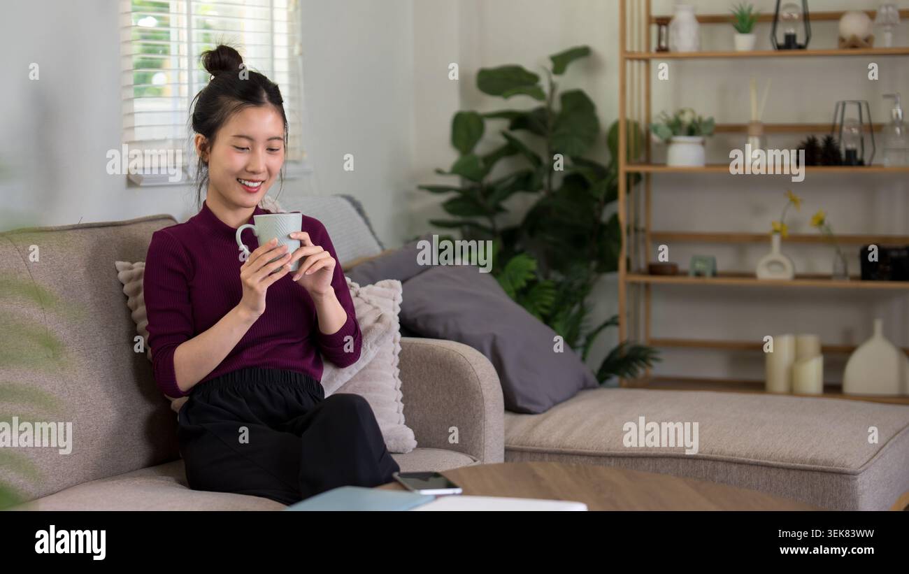 young Asian woman holding coffee mug relaxing on sofa smiling at home ...