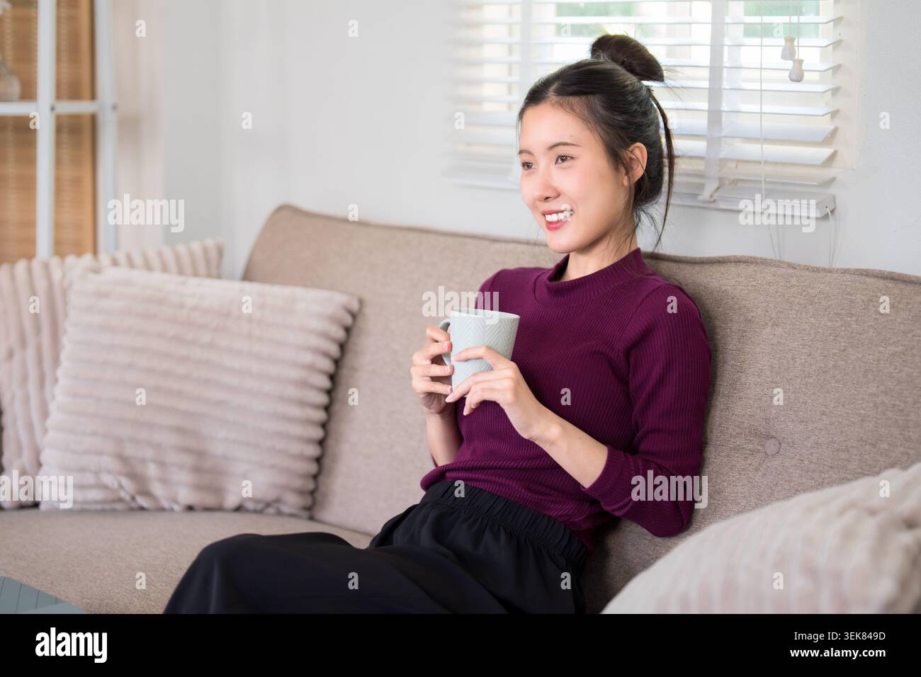 Asian women sitting on couch with coffee cup smiling in bright modern ...