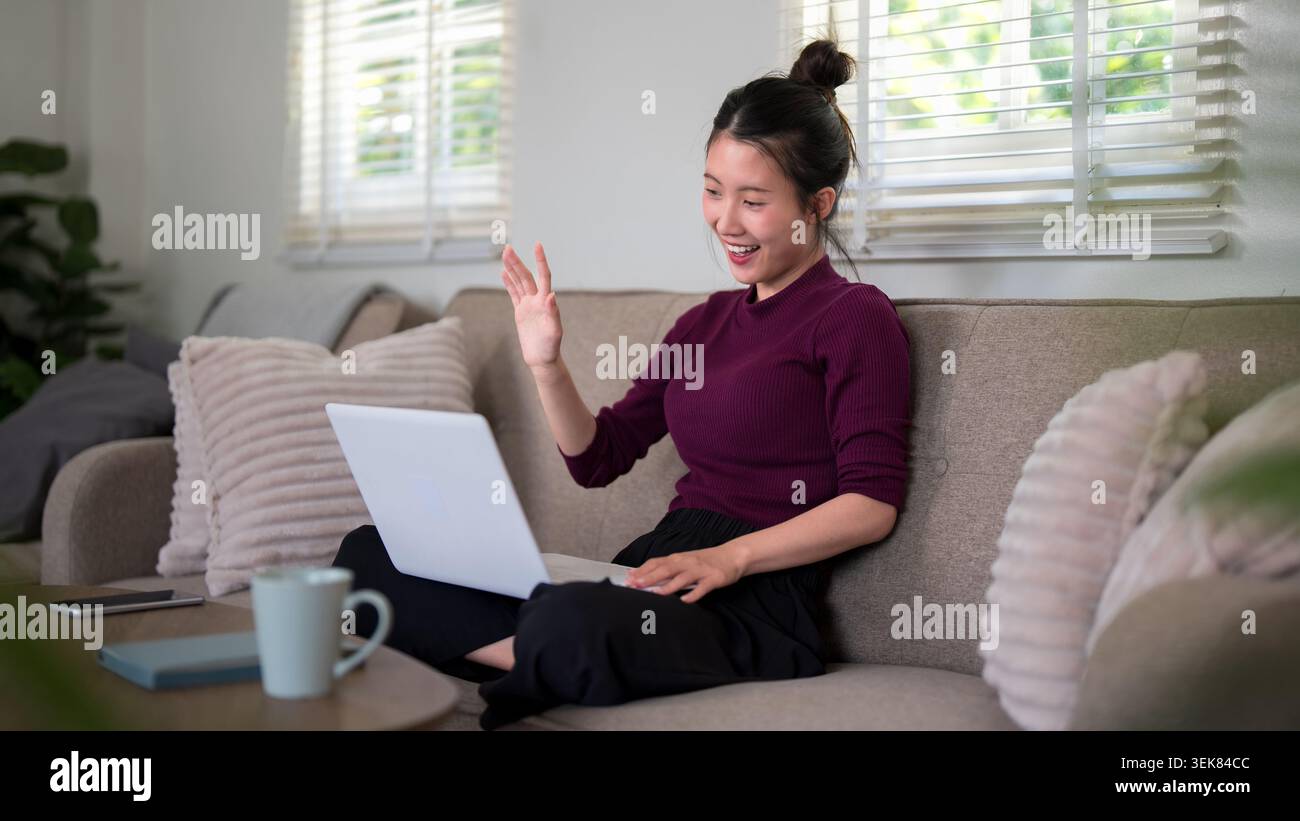 Young Asian woman sitting on couch waving at laptop during video call ...