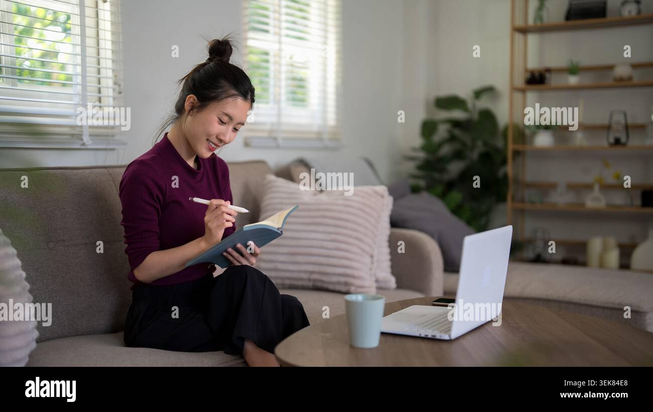 Asian woman writing notes in journal with laptop computer at home ...