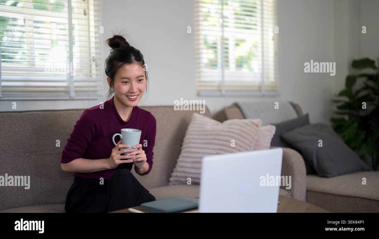 Asian woman holding coffee mug smiling at laptop computer working from ...