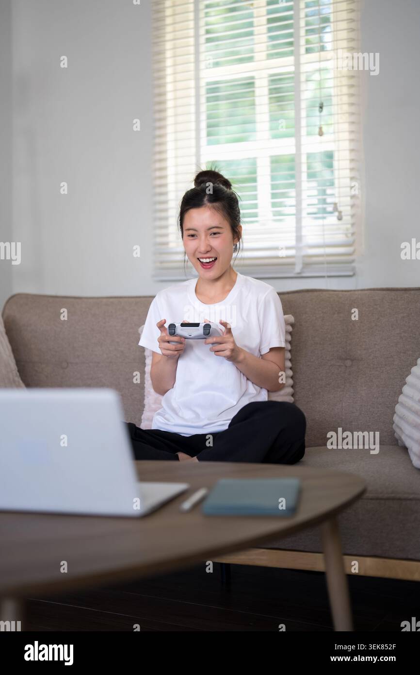 young Asian woman playing video game with controller sitting on sofa at ...