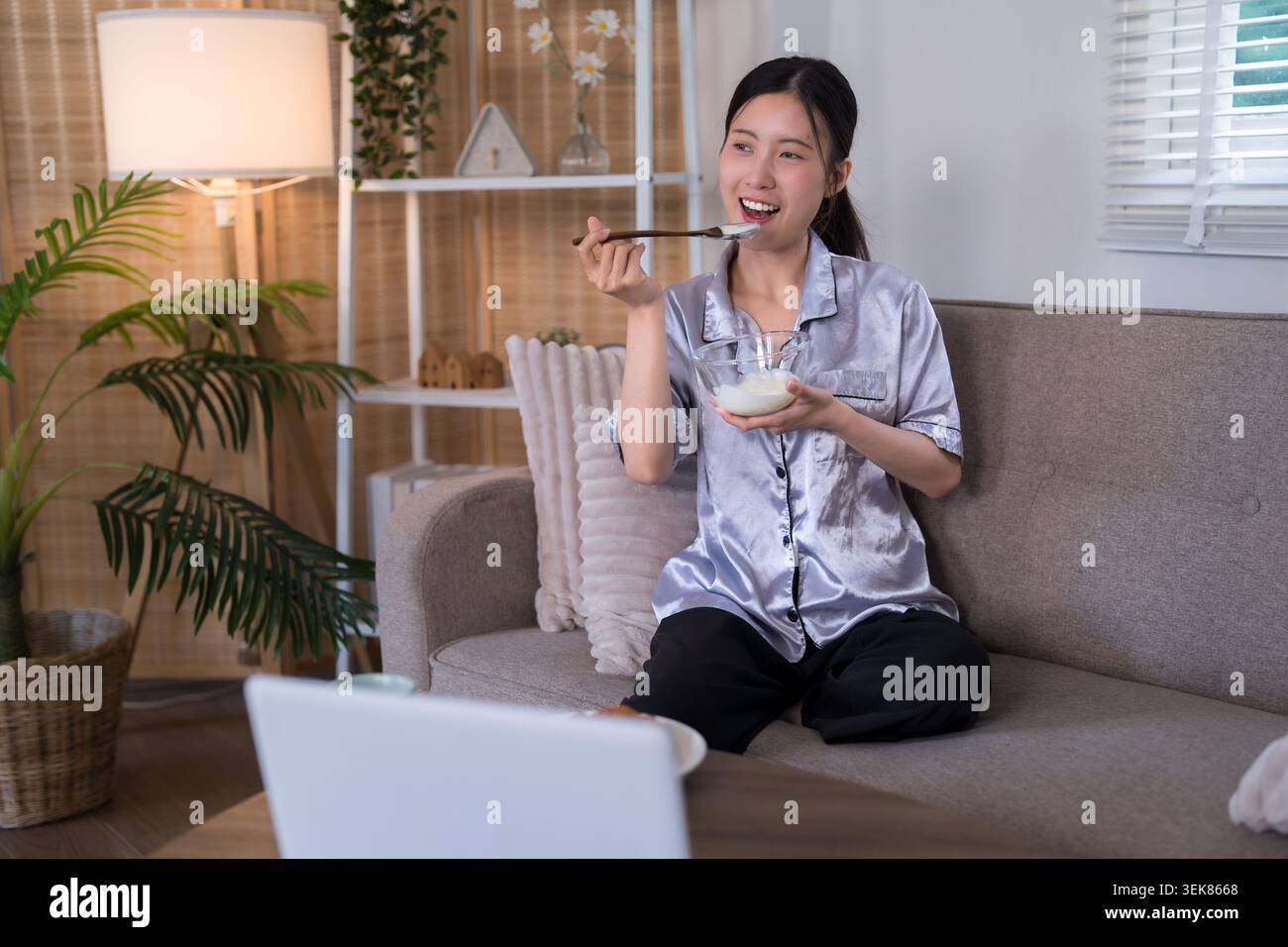 Asian women eating cereal breakfast bowl sitting on living room couch ...