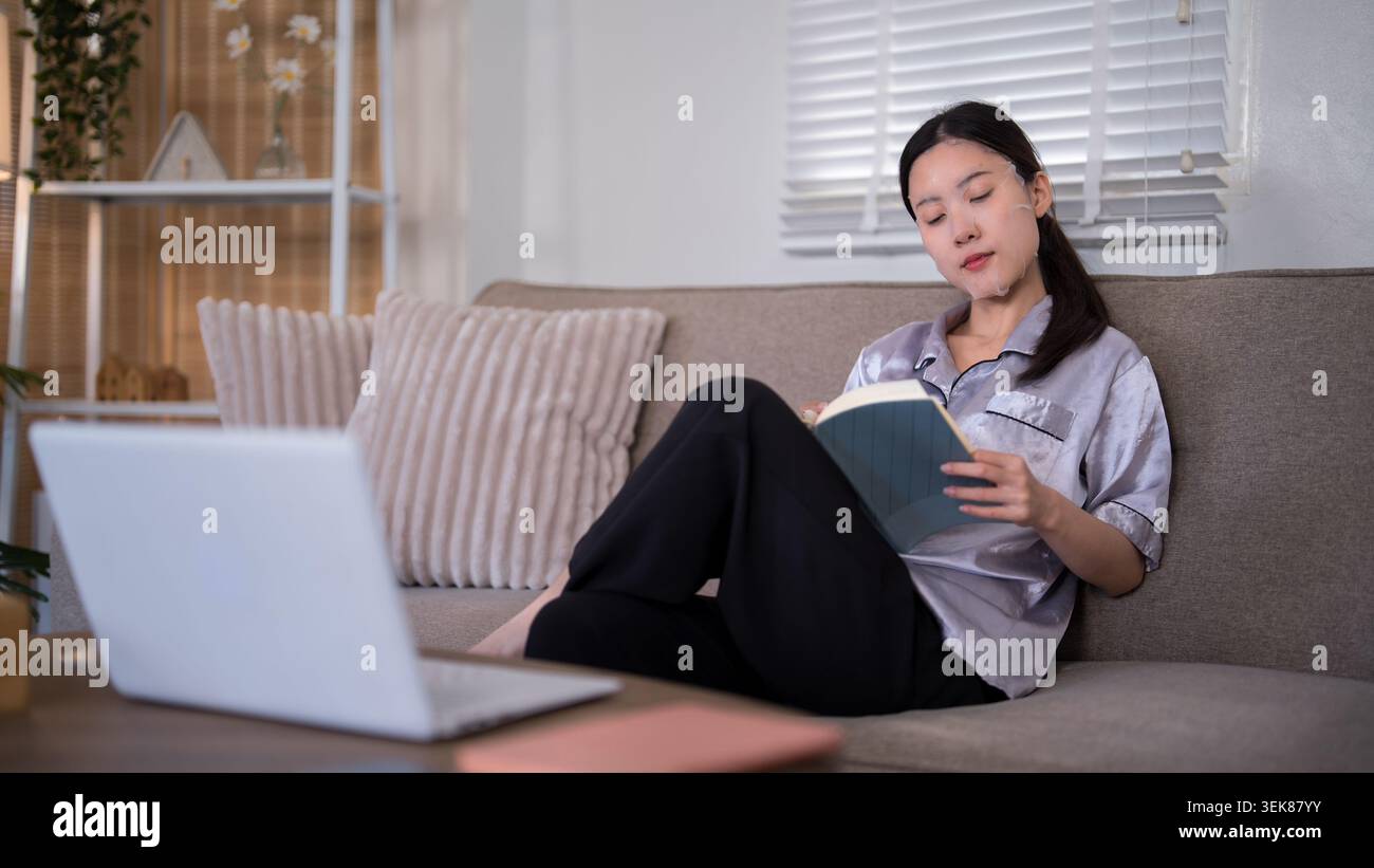 Asian woman doctor reading book resting on couch with laptop computer ...