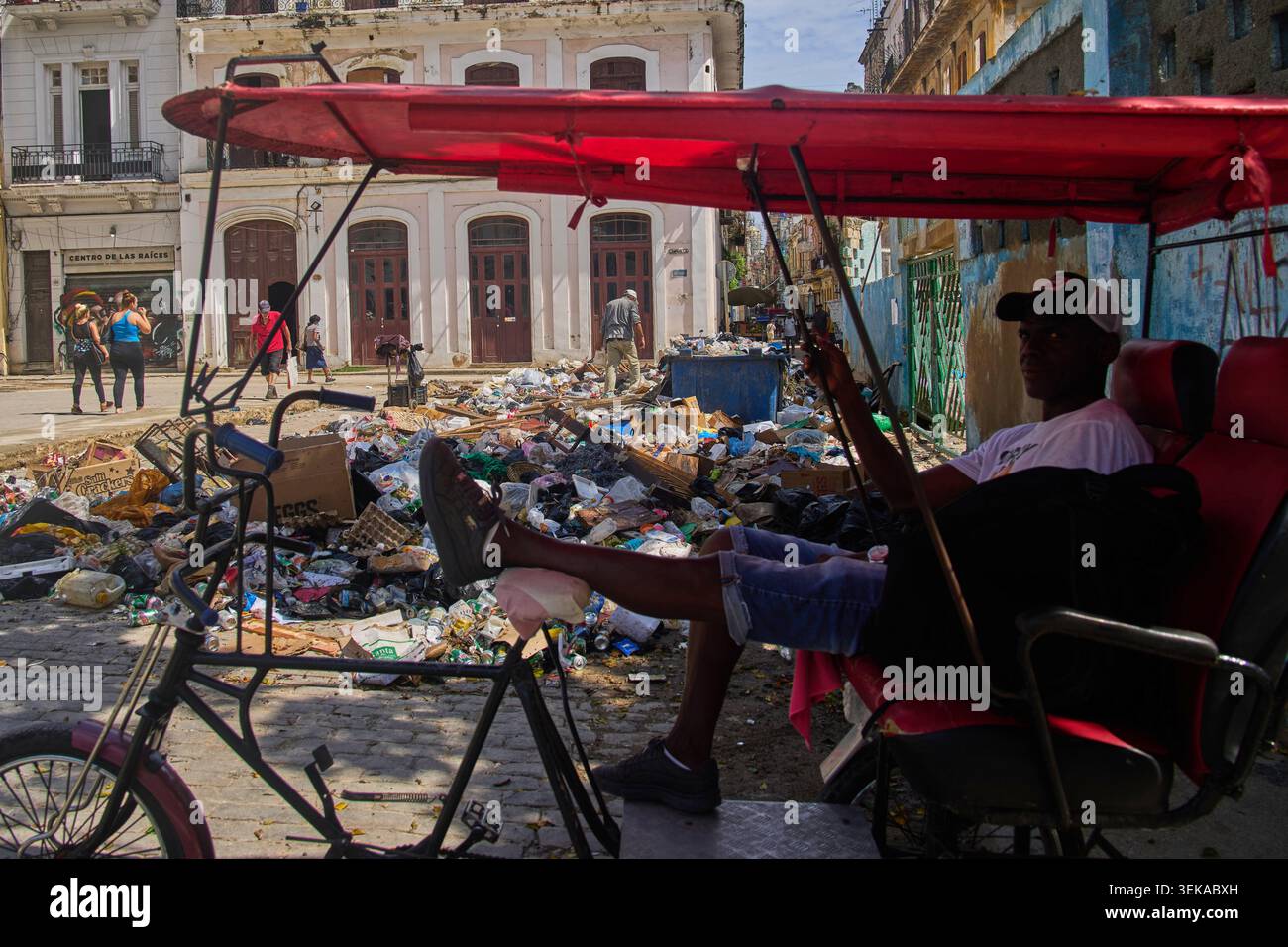 A bicycle taxi driver waits for customers, next to a pile of trash in ...