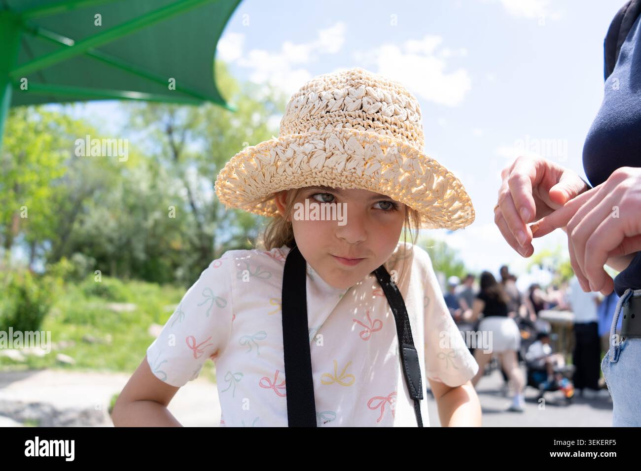 A young girl in a straw hat and patterned shirt stands outdoors under a ...