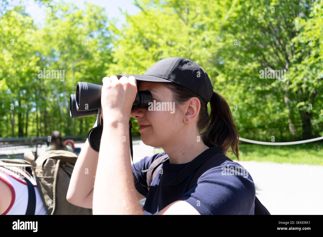 A young woman wearing a black cap and navy shirt uses binoculars to ...