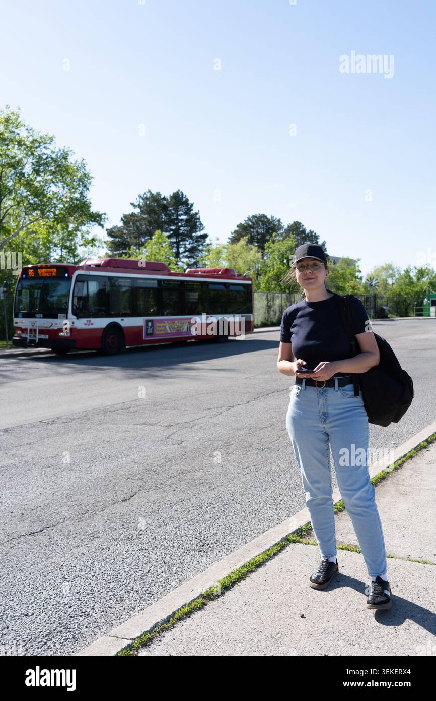 A person in casual clothing stands on a sidewalk holding a phone, with ...