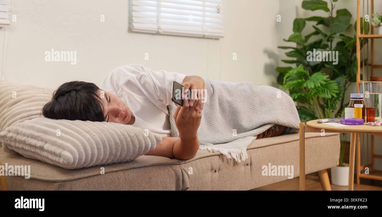 Asian man resting on couch using smartphone while sick at home Stock ...