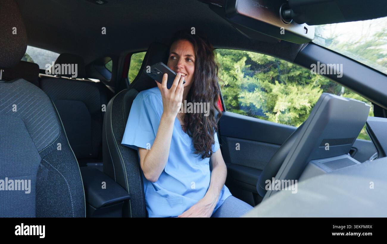 Woman in scrubs sending a voice message with smartphone inside a car ...