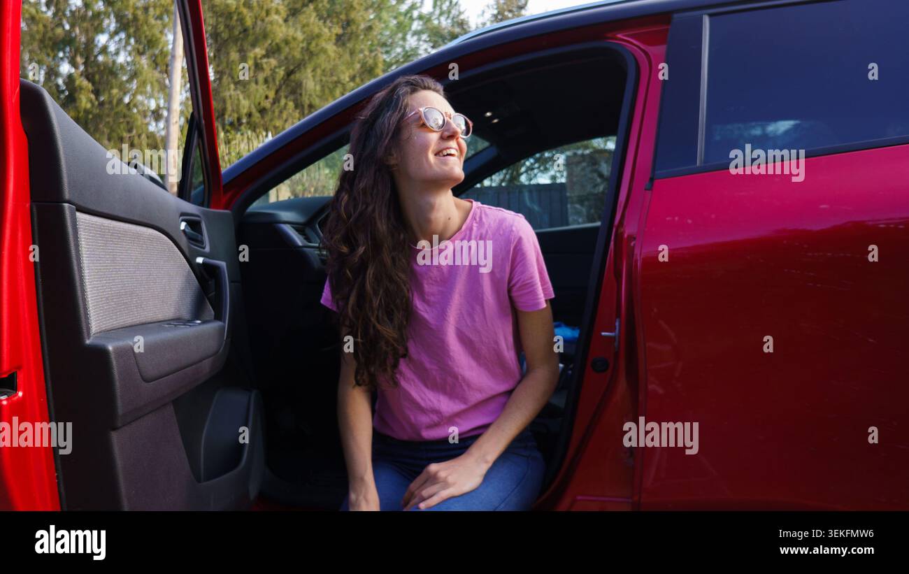 Young woman with sunglasses smiling, looking up while exiting red car ...