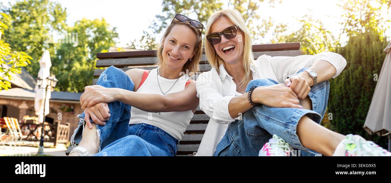Two smiling women sit on wooden bench sunny park enjoy relax moment ...